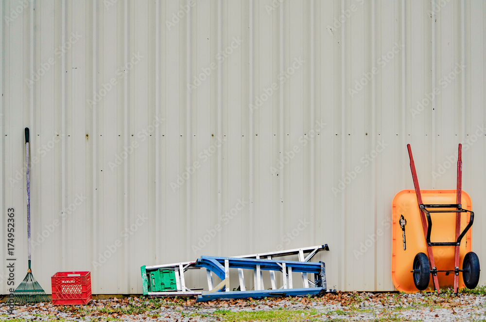 A rake, a milk crate, two ladders, and a wheelbarrow at the side of a ...