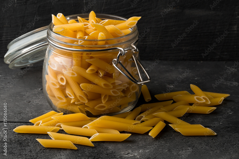 Raw, uncooked, dry penne pasta noodles in glass jar on black table ...