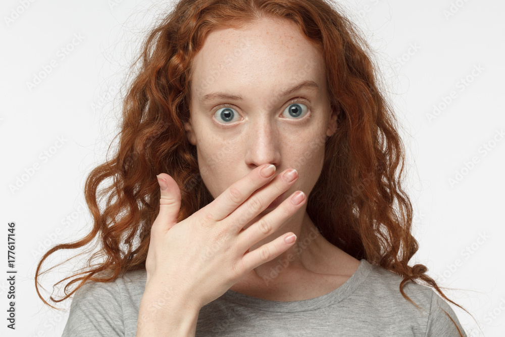 Astonished bug-eyed curly redhead girl covering her mouth with one hand ...