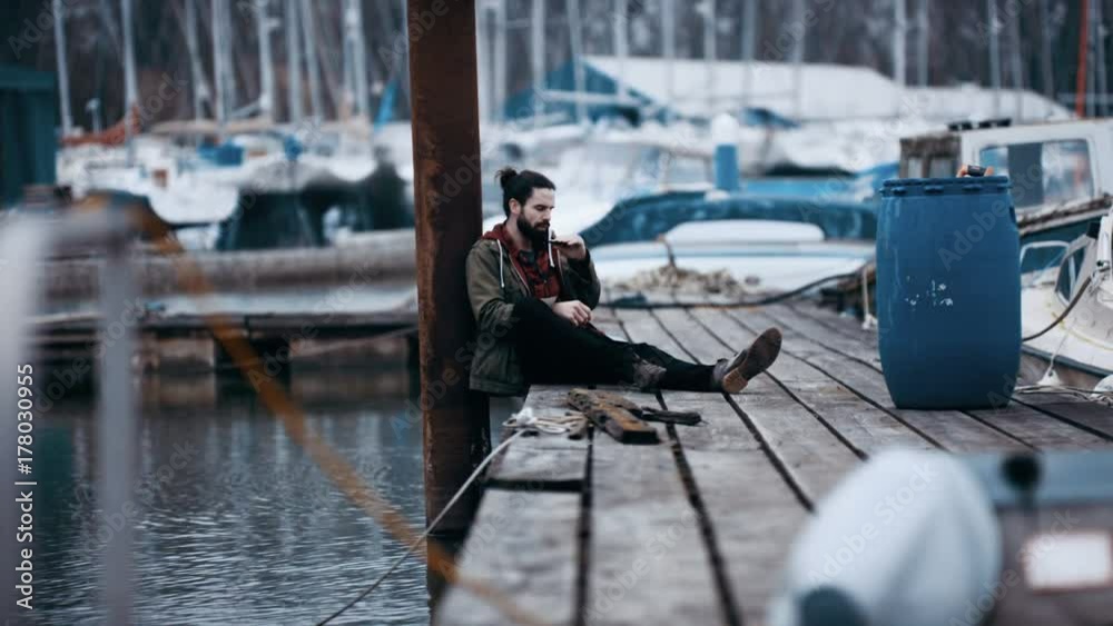 Rugged hipster man sitting alone on jetty in marina & playing harmonica ...