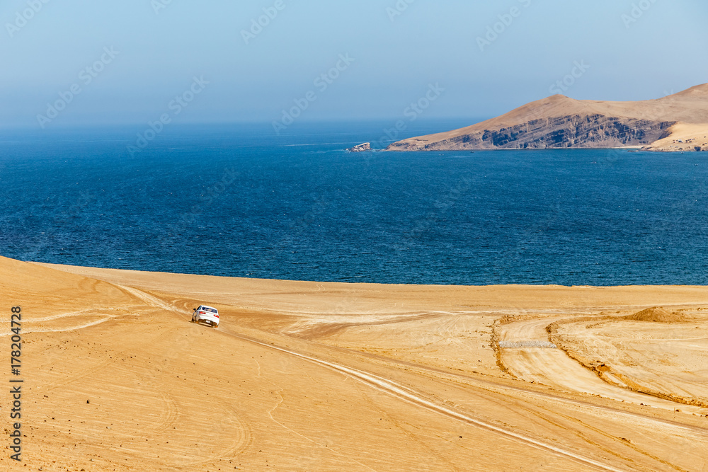 White car driving through Paracas desert toward the blue waters of ...