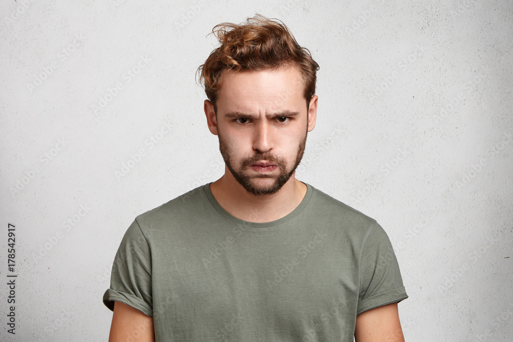 Portrait of sad bearded young man has grumpy expression pouts his lips ...