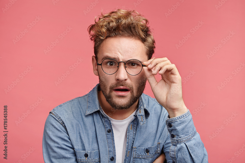 Scrupulous stylish man in denim shirt and spectacles looks with shocked ...