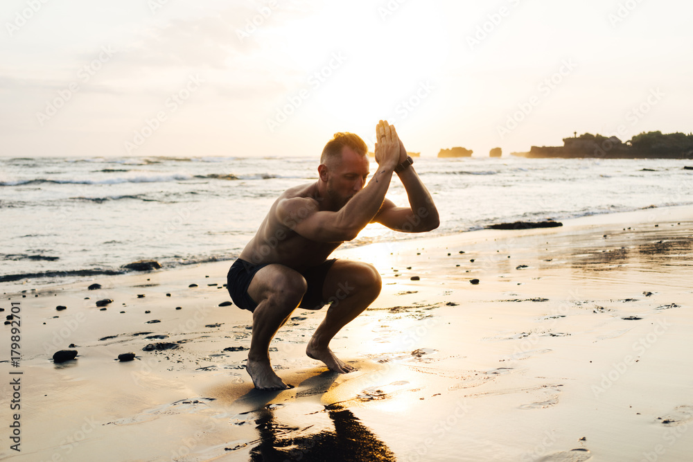 Handsome bearded muscular male is doing squat exercises on a wild sand ...