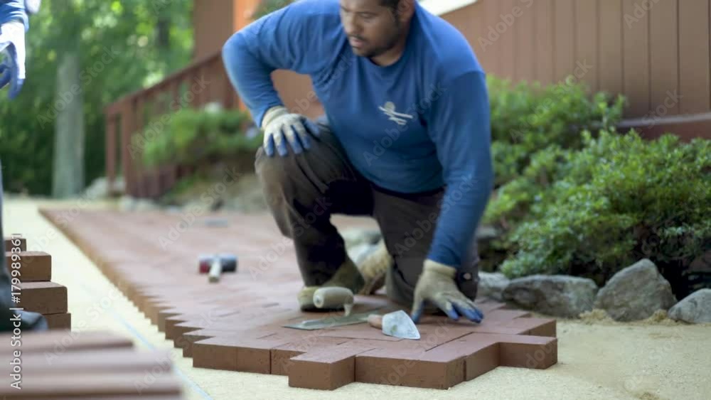 One man hands red bricks to another man who is putting the brick pavers ...