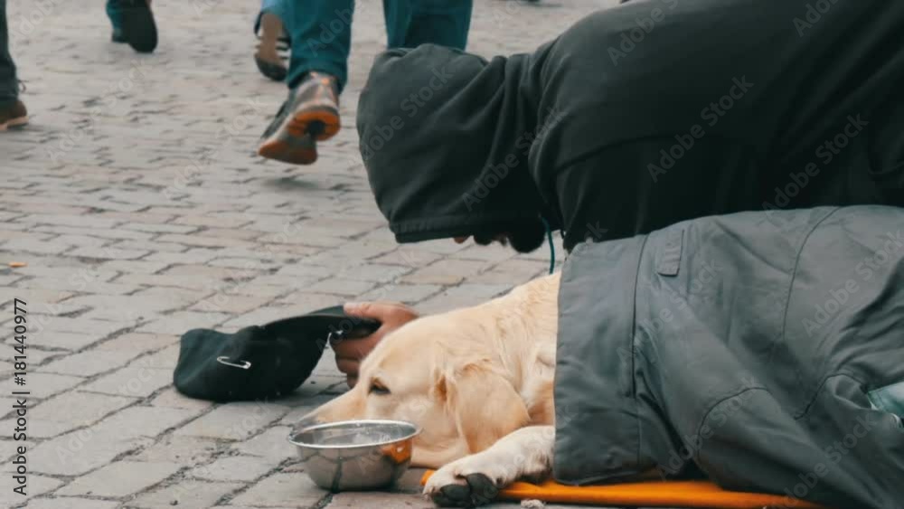 Poor man kneels and begs alms with his outstretched hand next to the lying dog. A beggar is ...