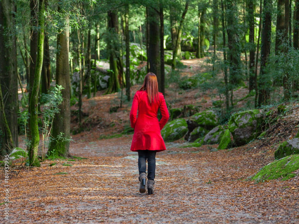 Young woman walking away alone on forest path wearing red long coat or ...