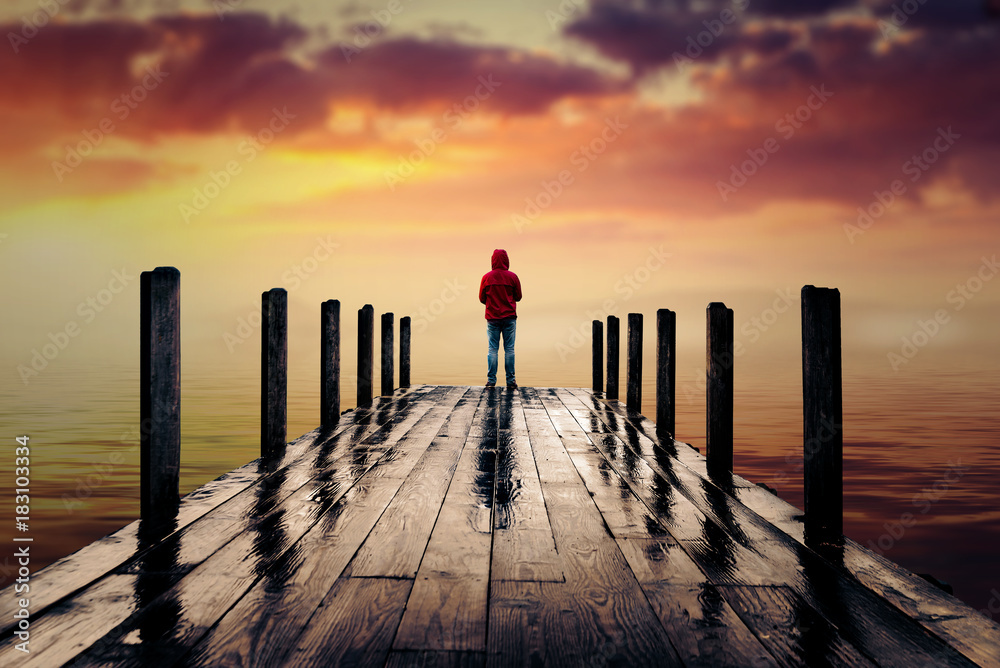 lonely man standing on a wooden pier during sunset facing mountain ...