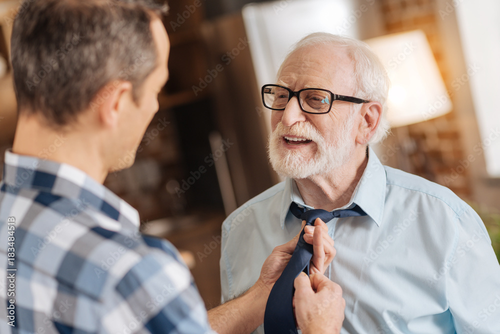 Vital help. Caring young man helping his elderly father tie his tie and ...