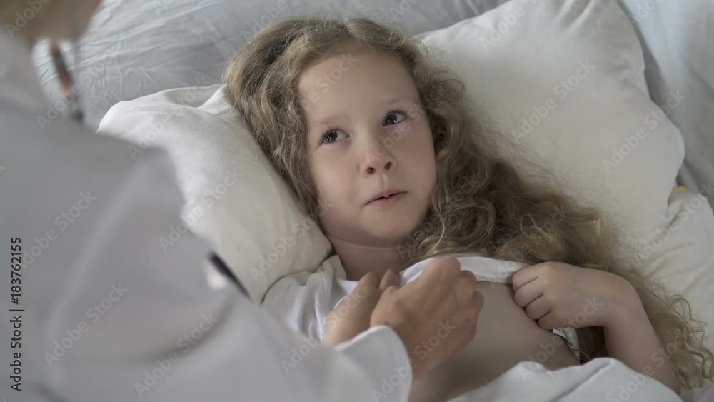 Sick kid lying in bed and crying while doctor examining her with ...