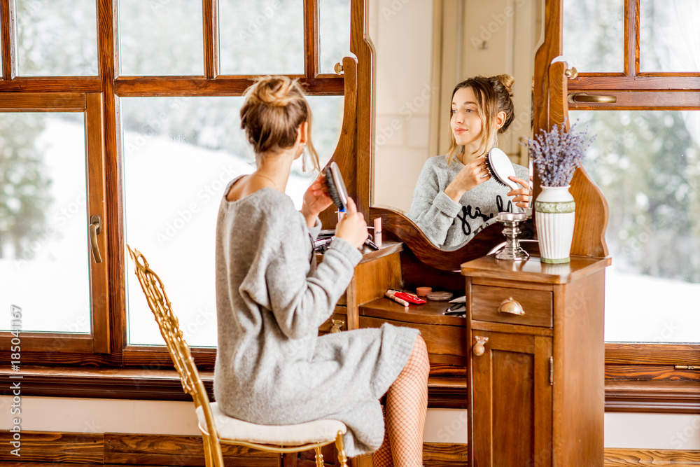 Woman brushing hair at the dressing table near the window at the wooden ...