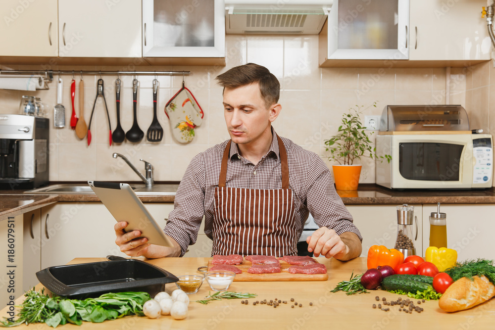 Young European man in apron sitting at table with vegetables, looking ...