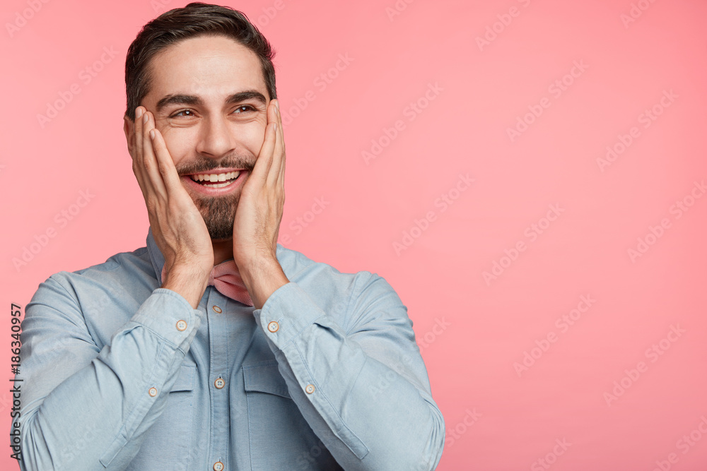 Content cheerful male feels shy and embarrasment, keeps hands on cheeks ...