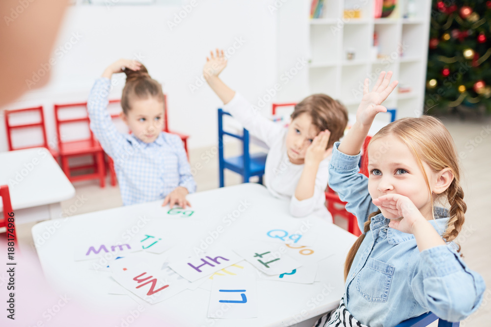 Cute kids by table raising their hands while learning alphabet with ...