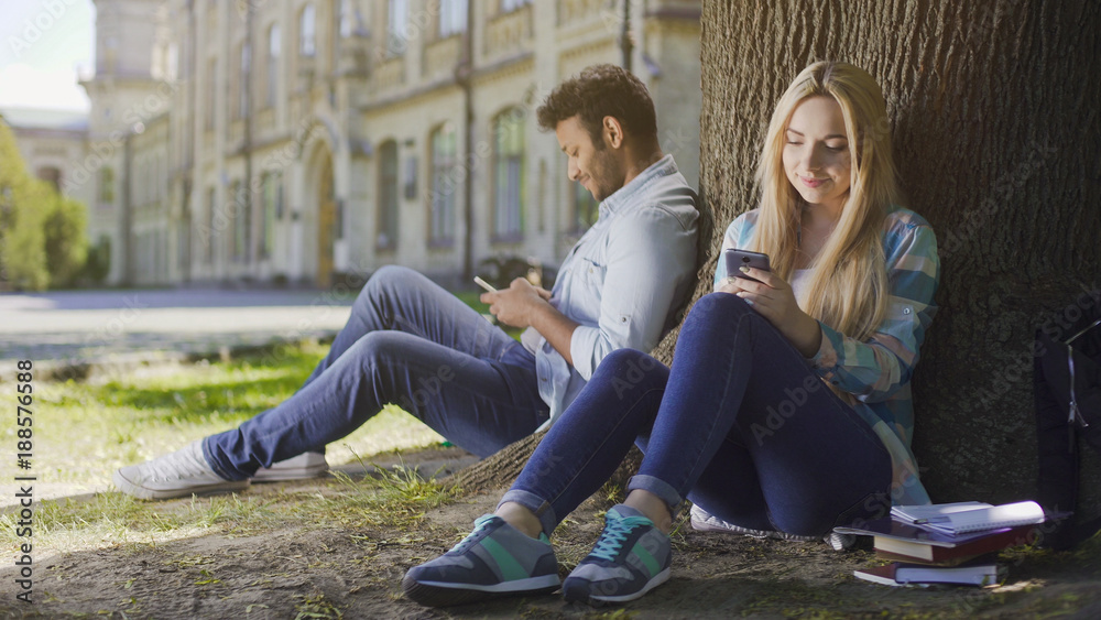 Multiracial guy and girl sitting under tree using mobile phones smiling ...