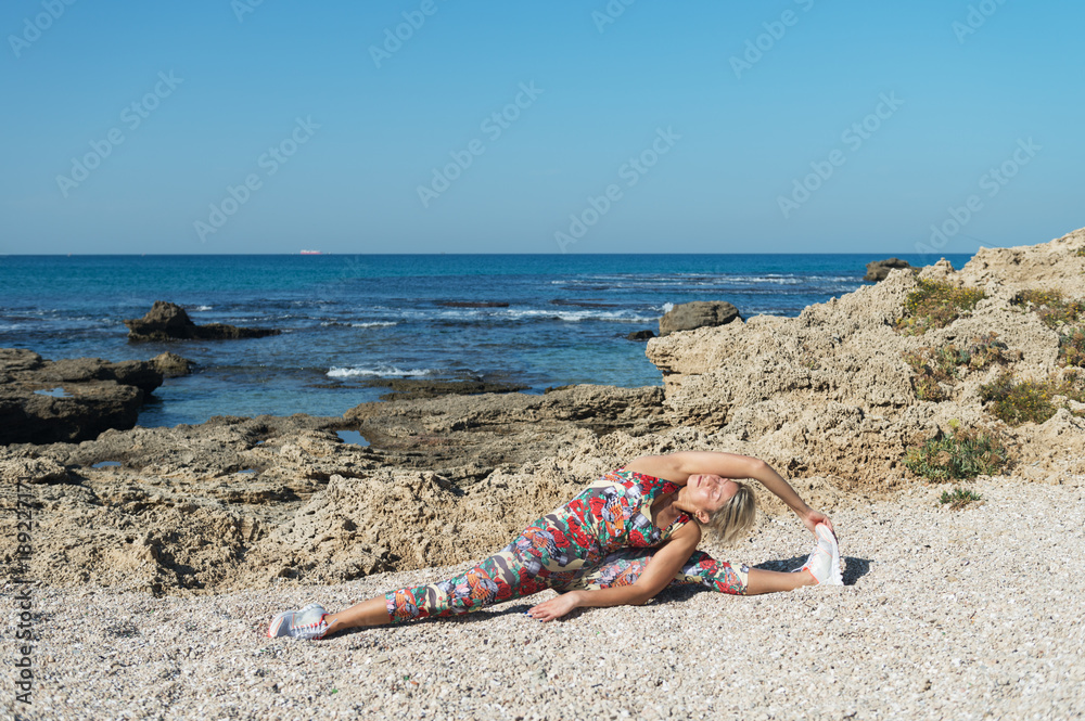 white blond female athlete making split on the beach with a sea at the ...