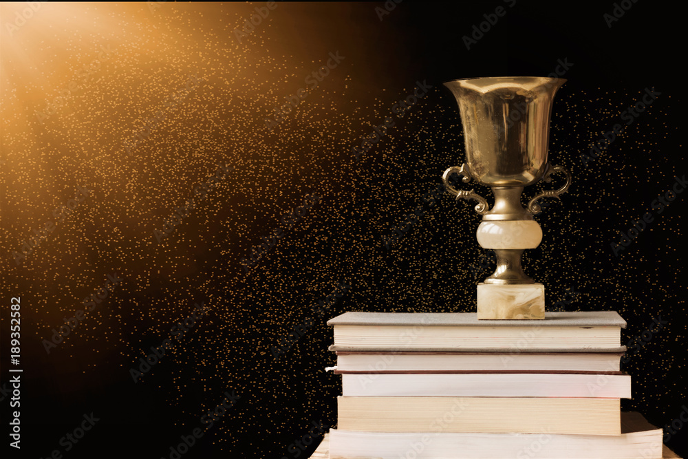 Golden trophy on pile of books, against blackboard, with sun rays over ...