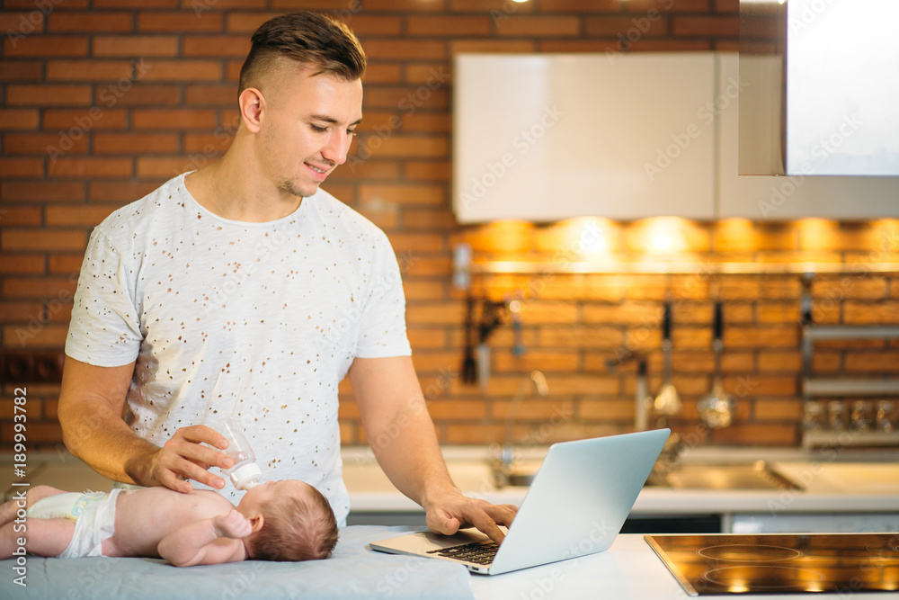 dad trying to work while standing with his newborn babe in home office ...