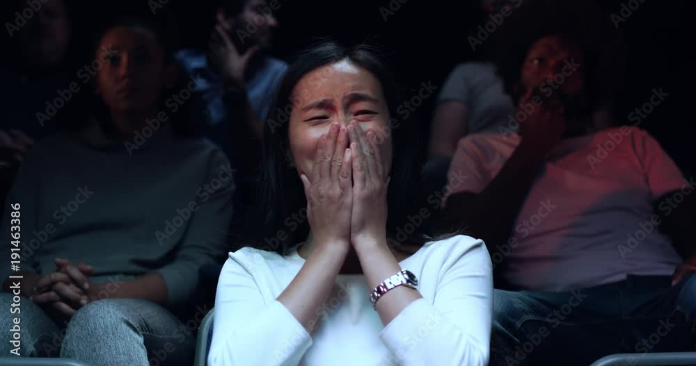 A Chinese woman in a movie theatre holds her hands to her face as she ...