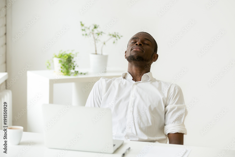 African office worker relaxing with eyes closed sitting at work desk ...