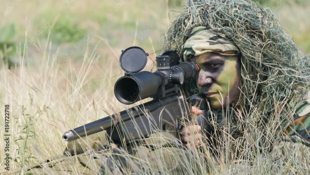 Army sniper wearing disguise camouflage suit lying and aiming in grass ...