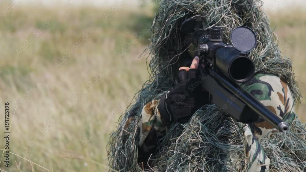 Army sniper wearing disguise camouflage suit sitting and aiming in ...
