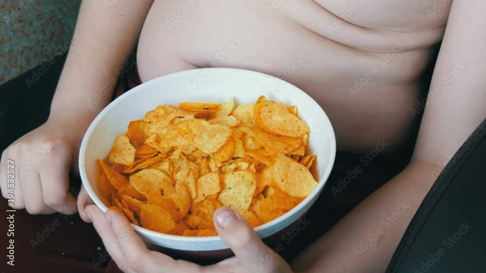 Fat teenager with fat folds on his body is eating potato chips ...