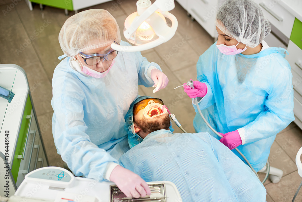 Two female dentists in blue robes are attending the teeth of a male ...