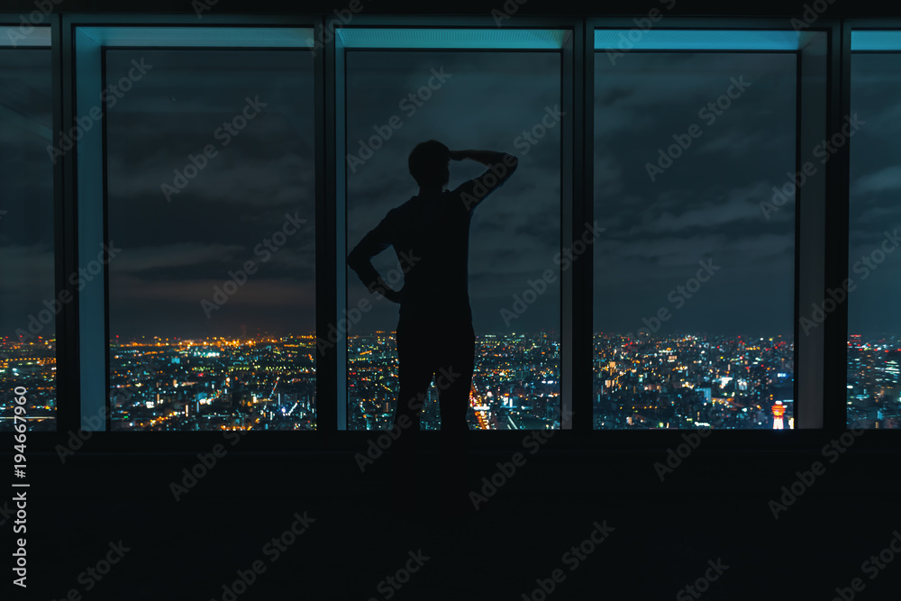 Man looking out large windows high above a sprawling city at night ...