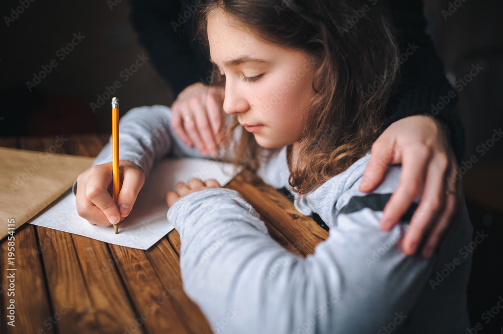 Little girl doing homework. The child writes a letter. Mom is standing ...