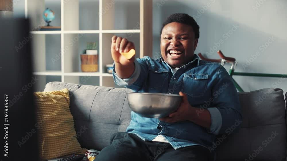 Happy rude african american man watching tv sit on couch eat chips ...
