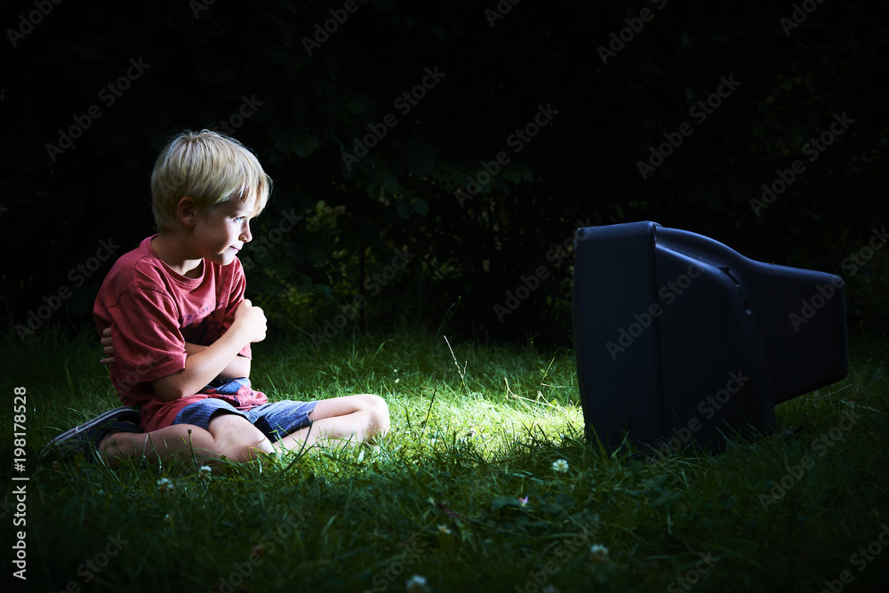 Child blond boy watching TV horror / thriller. Outside in the garden at ...