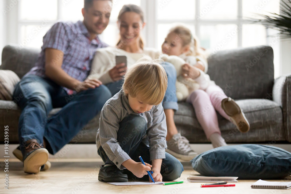 Serious cute son holding color marker drawing on floor while parents ...