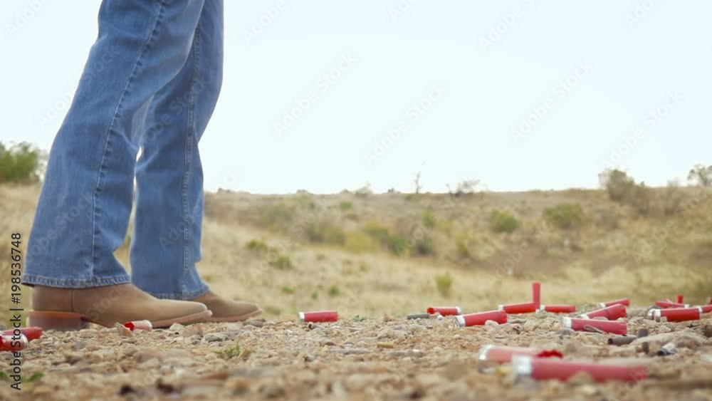 Low angle of a man in cowboy boots shooting a shotgun. Red cartridges ...