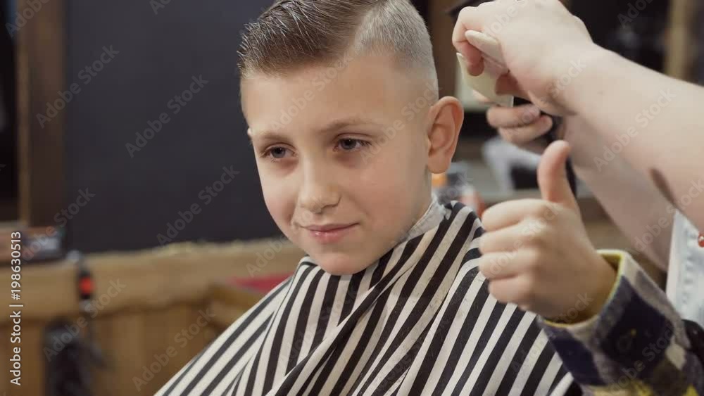 A cute boy sits on a chair in a barbershop look in the camera and shows ...