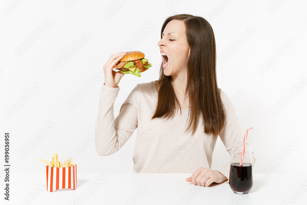 Hungry young woman greedily eats burger, sits at table with french ...