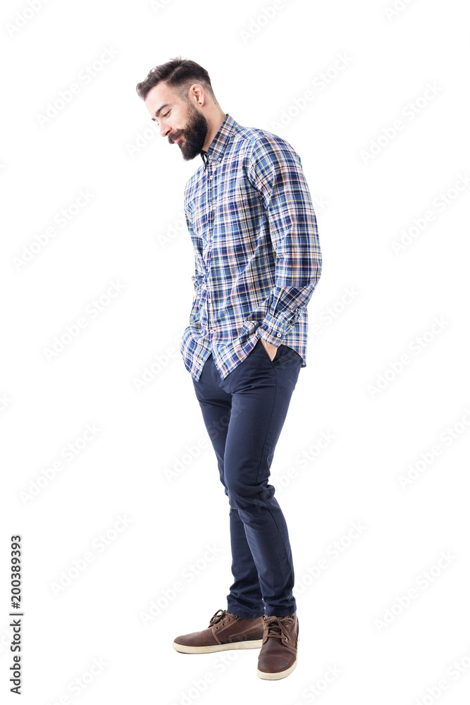Shy handsome bearded young man in plaid shirt with hands in pockets ...