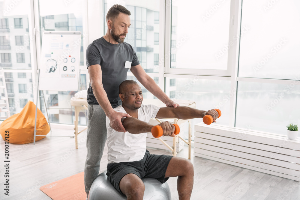 Developing strength. Pleasant young man looking at the dumbbells while ...