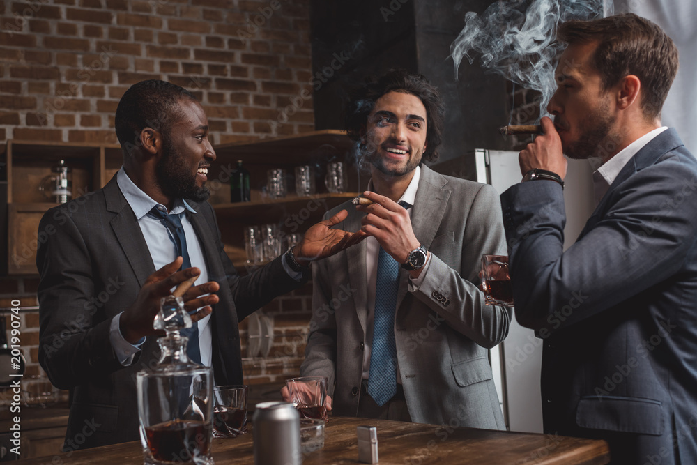 smiling multiethnic male friends in suits talking, drinking whiskey and ...