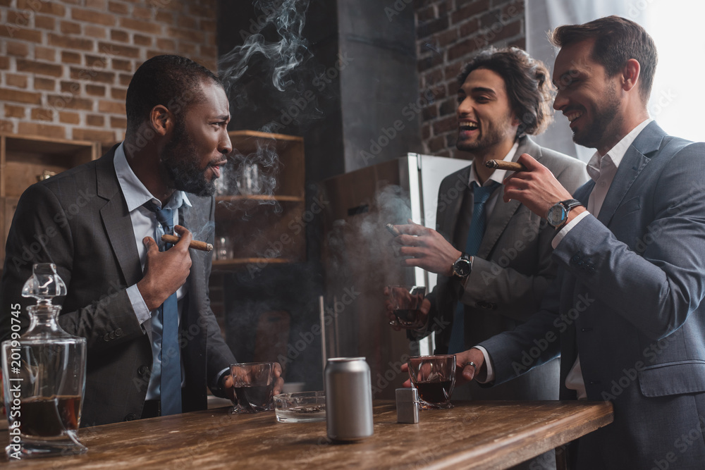 smiling multiethnic male friends in suits smoking cigars, drinking ...