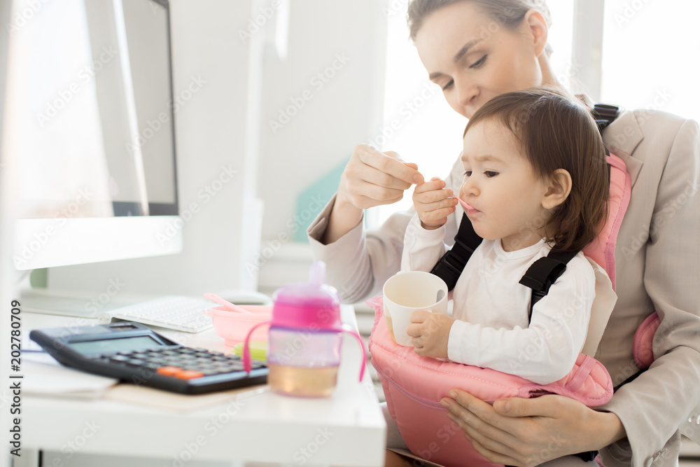 Young office worker feeding her baby daughter in carrier with special ...