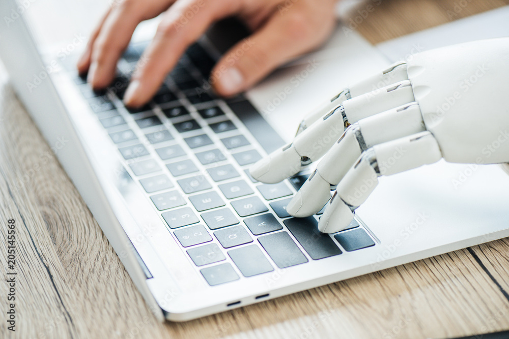 close-up view of human and robot hands typing on laptop at workplace ...