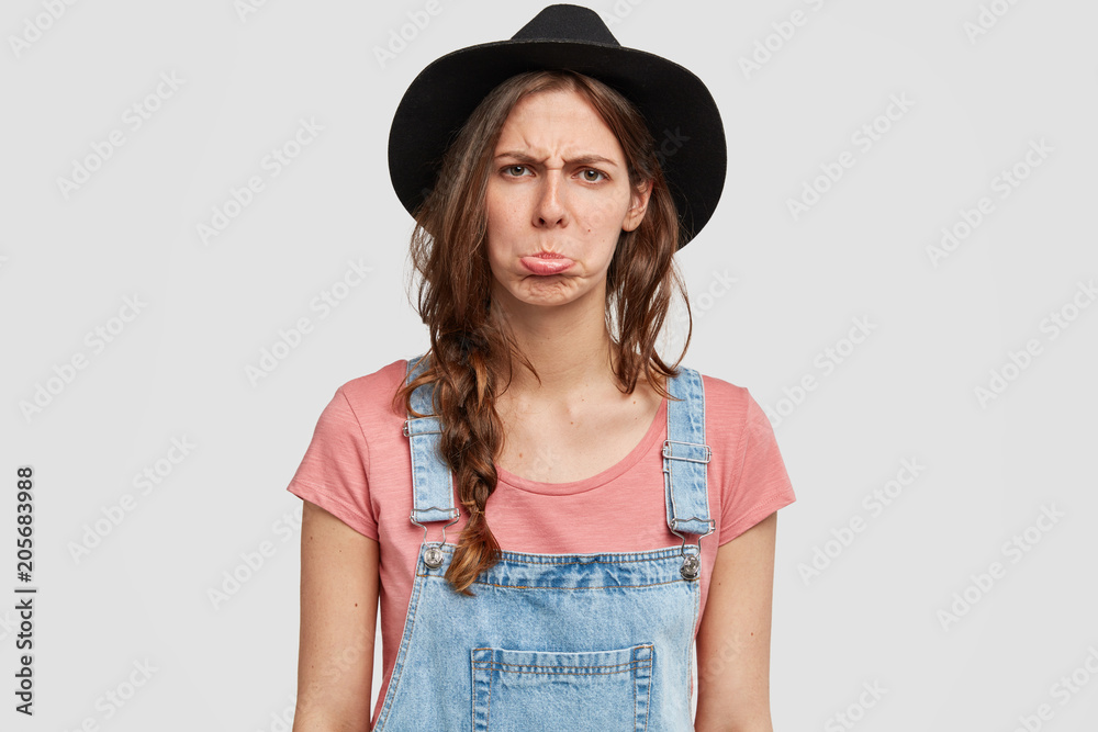 Displeased young female curves lower lip, wears black hat, denim ...