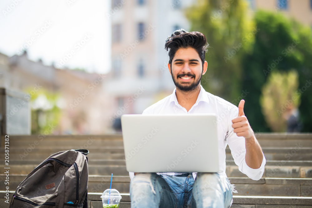 Happy indian student sitting on the stairs showing thumb up working on ...