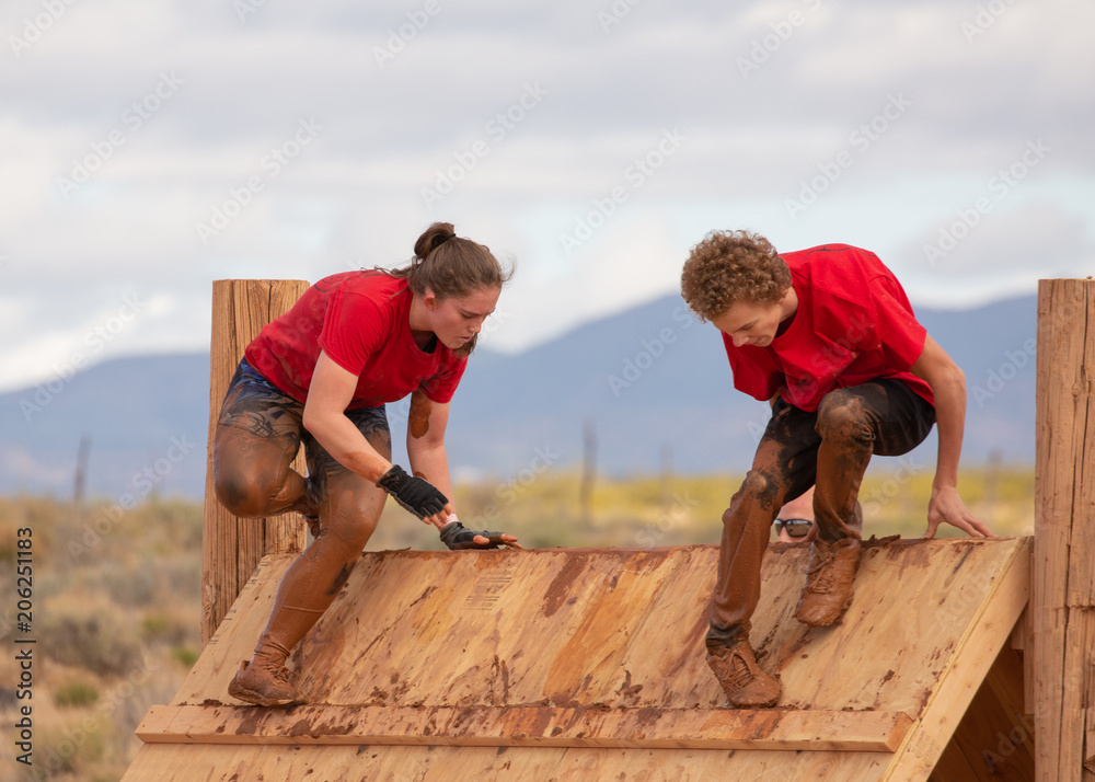 Teenage boy and girl climbing over an obstacle in a mud run obstacle ...