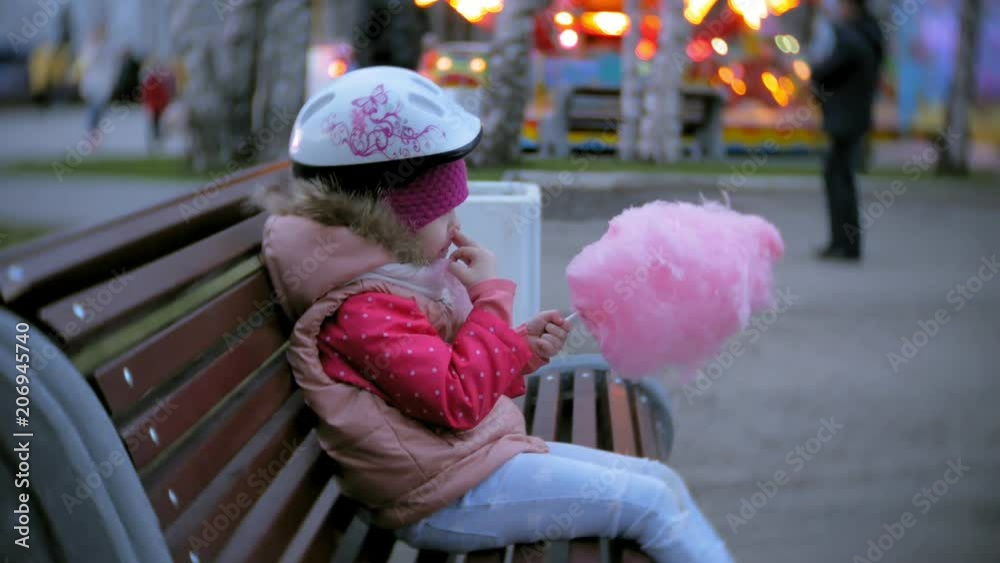 Beautiful little girl sitting on a bench in an amusement park, eating ...