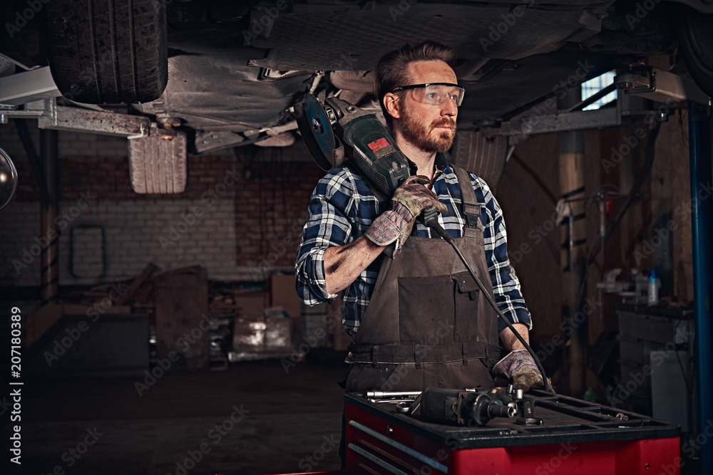Mechanic in a uniform and safety glasses holds an angle grinder while ...