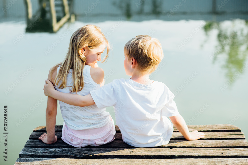 Back view of two blond children sitting together on pier of wooden ...