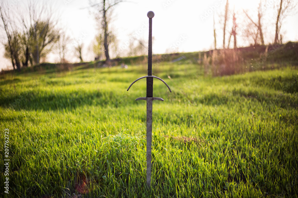 two-handed sword in the ground, against the background of a green field ...