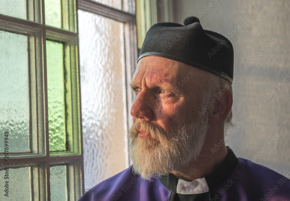 Mature priest looking out of a church window with a thoughtful ...